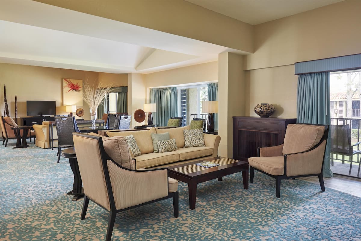 Quarto de hotel com sofá, poltronas, mesa de centro, televisão e uma janela grande com cortinas.
