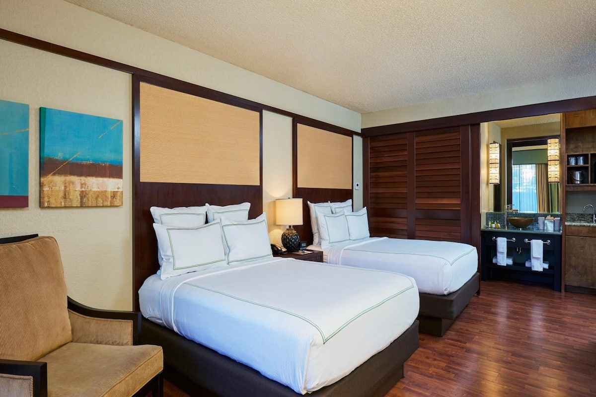 Quarto de hotel com duas camas, uma cadeira, uma mesa de trabalho com abajur e um banheiro visível através de uma porta aberta.