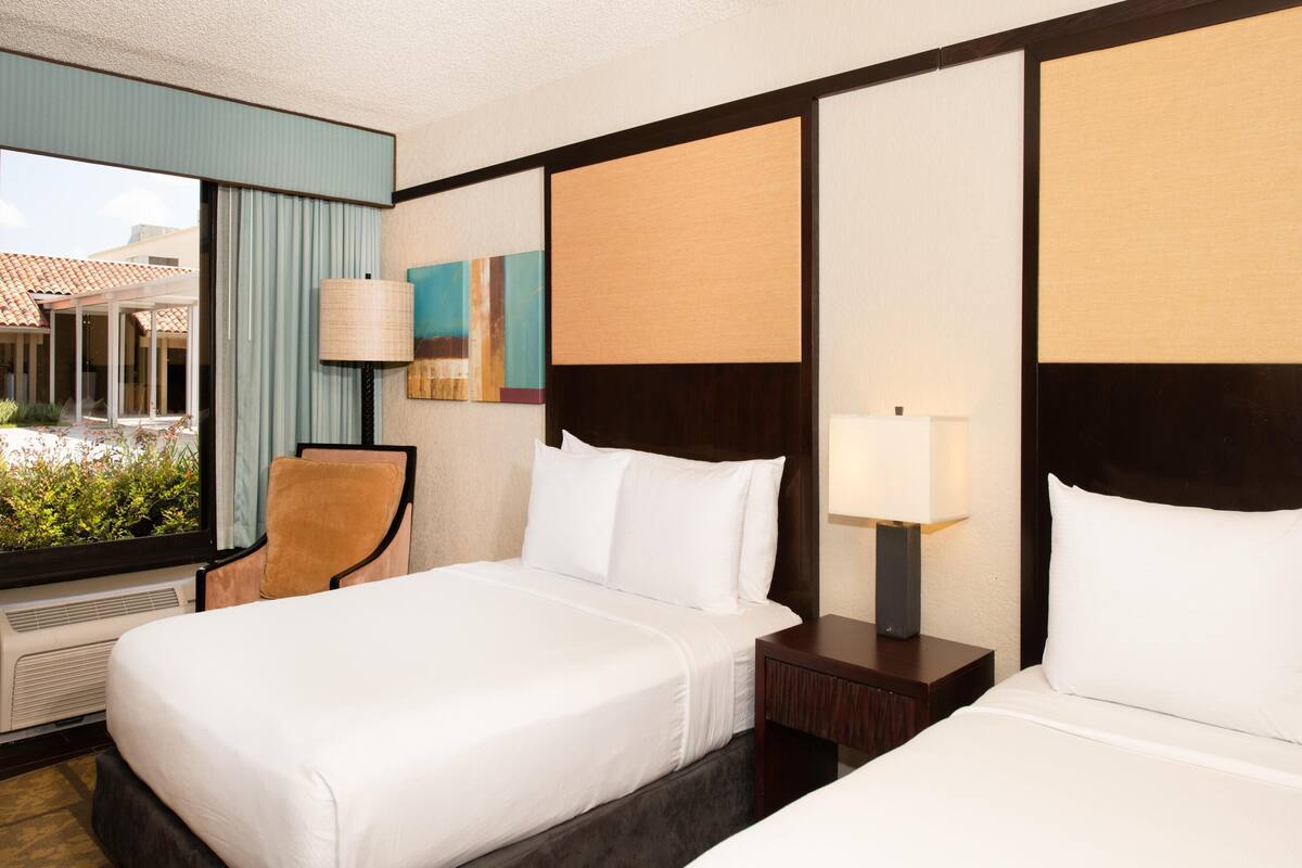 Quarto de hotel com cama, poltrona, mesa de cabeceira, luminária e janela com vista para edifícios e áreas verdes.