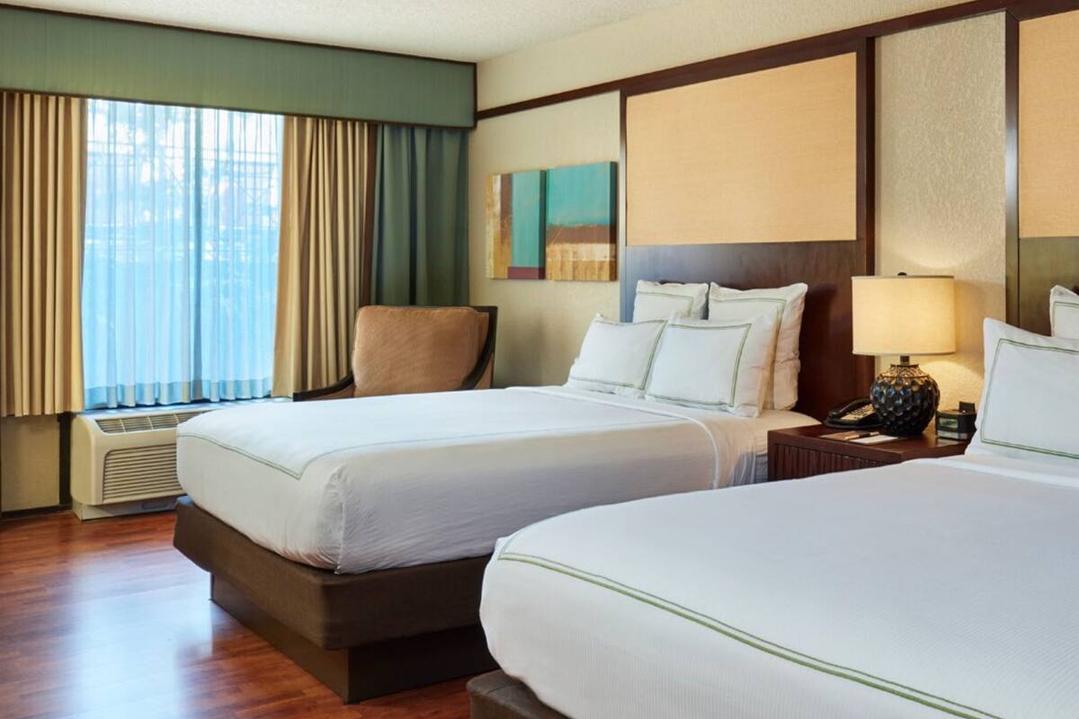 Quarto de hotel com duas camas, uma cadeira, uma escrivaninha com abajur e uma janela com cortinas.
