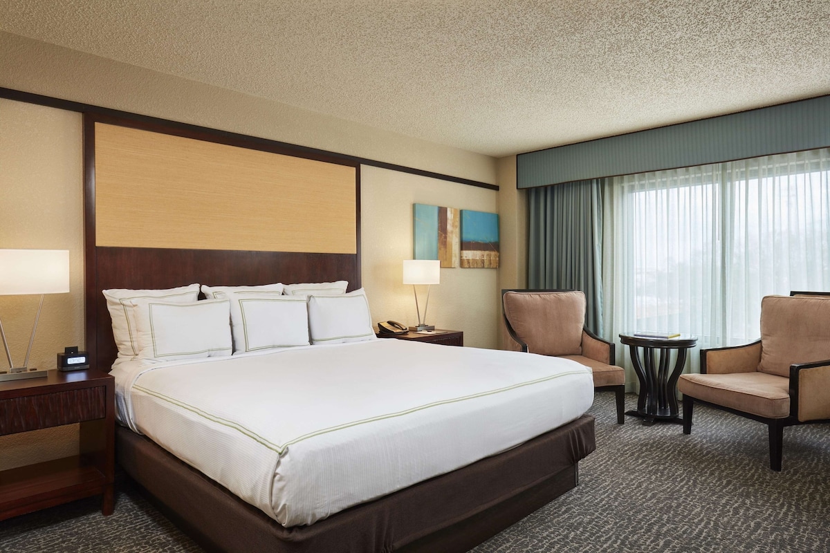 Quarto de hotel com uma cama grande, duas poltronas, uma mesa pequena e uma janela com cortinas.