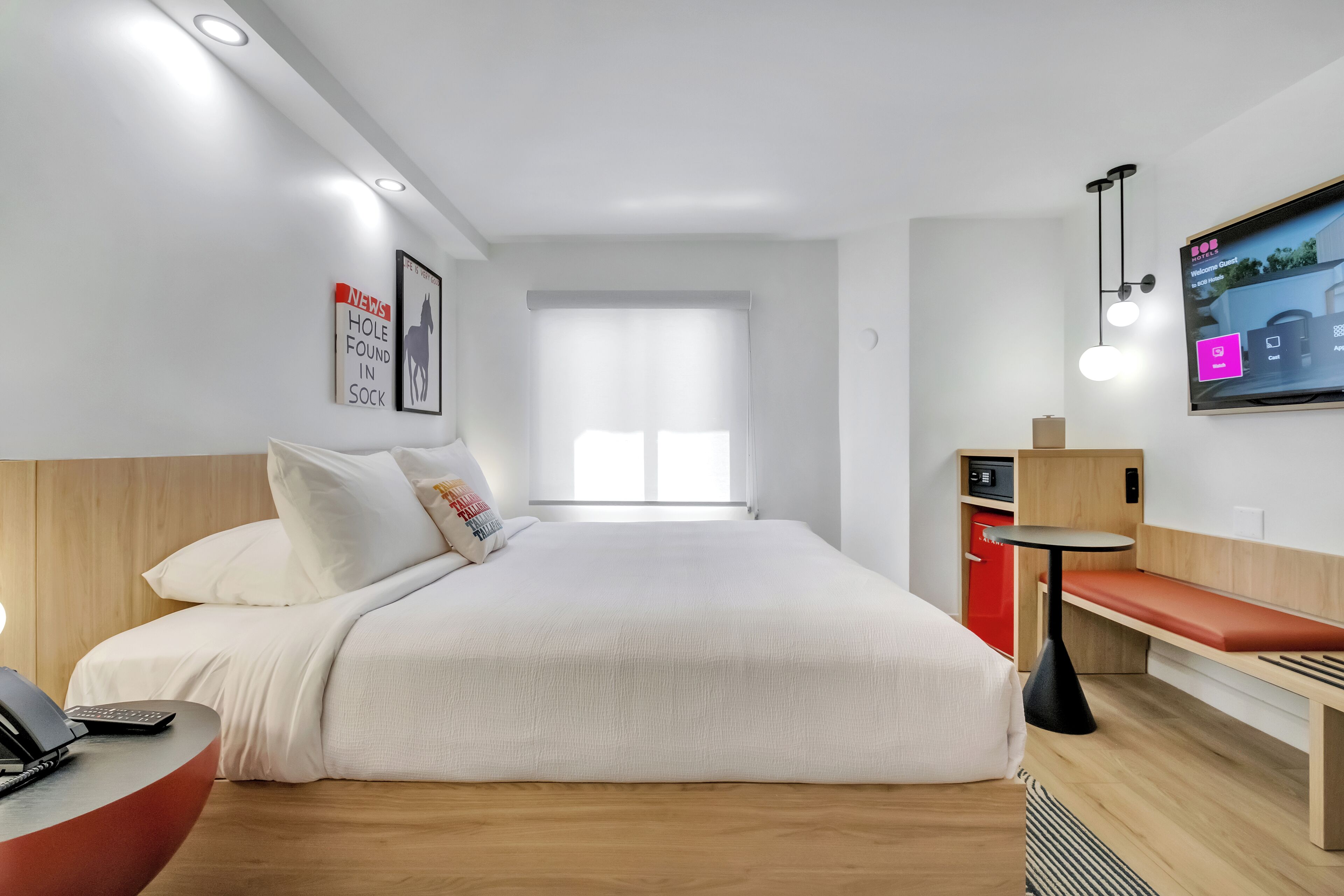 Quarto de hotel moderno com uma cama grande, uma mesinha de cabeceira com telefone, uma pequena escrivaninha com cadeira, uma TV na parede e uma janela com persianas.