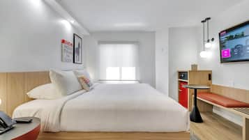 Quarto de hotel moderno com uma cama grande, uma mesinha de cabeceira com telefone, uma pequena escrivaninha com cadeira, uma TV na parede e uma janela com persianas.