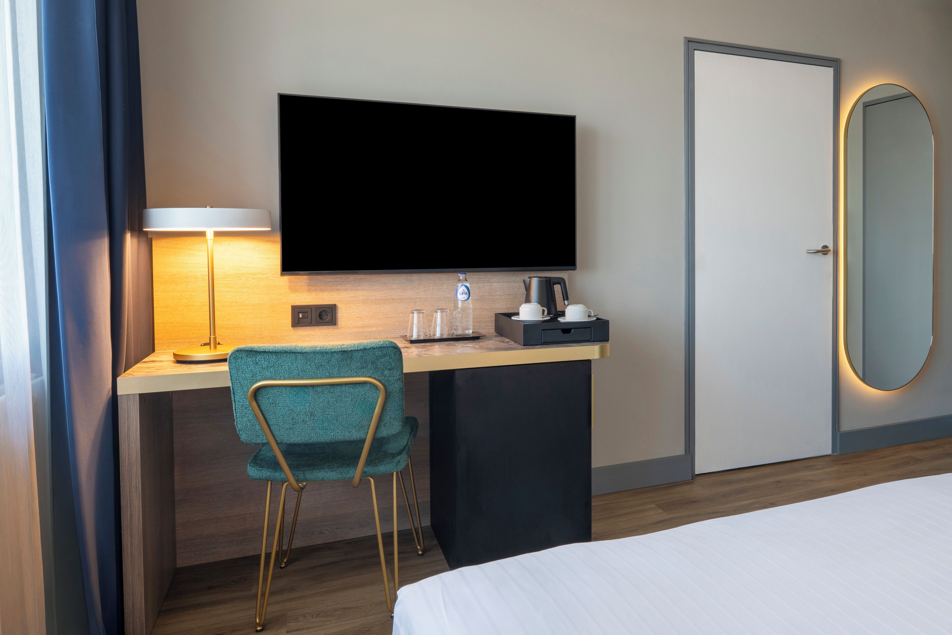 Quarto de hotel com cama, escrivaninha com cadeira, televisão de tela plana, cafeteira, espelho e porta.