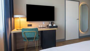 Quarto de hotel com cama, escrivaninha com cadeira, televisão de tela plana, cafeteira, espelho e porta.