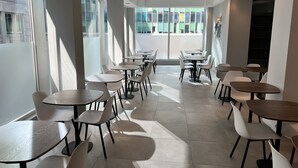 Interior moderno de café com várias mesas e cadeiras, grandes janelas e vista para a rua.