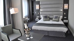 Quarto de hotel moderno com uma cama grande, uma mesinha de cabeceira com luminária, uma cadeira e uma pequena mesa com um vaso e dois copos.