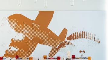 Restaurante com mesas montadas para refeições, um mural grande na parede e luminárias pendentes.