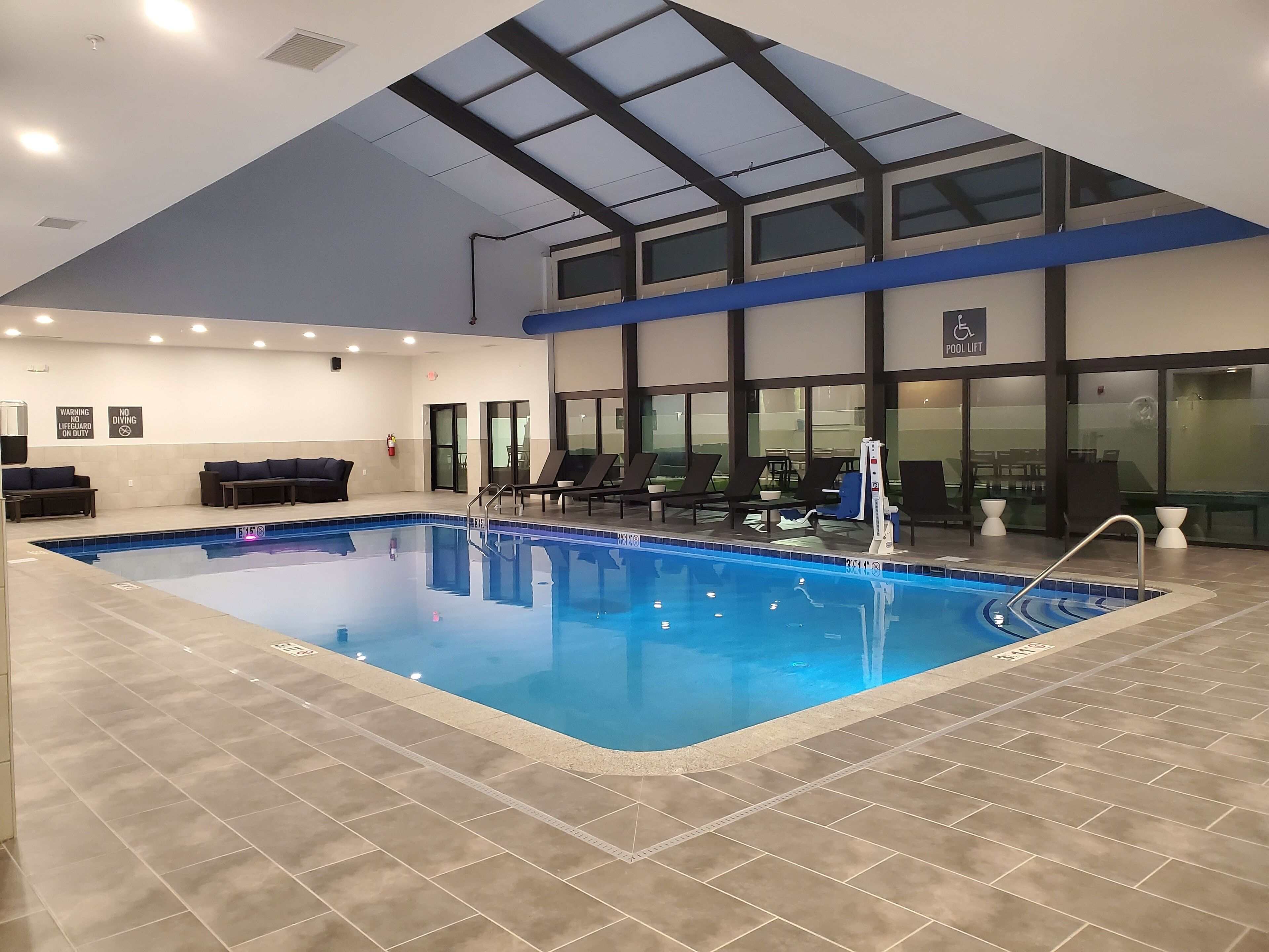 Piscina interior com chão em mosaico, cadeiras de descanso e teto com claraboias.