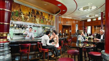 Uma área de bar com vários bancos e mesas, um barman e um mural na parede.