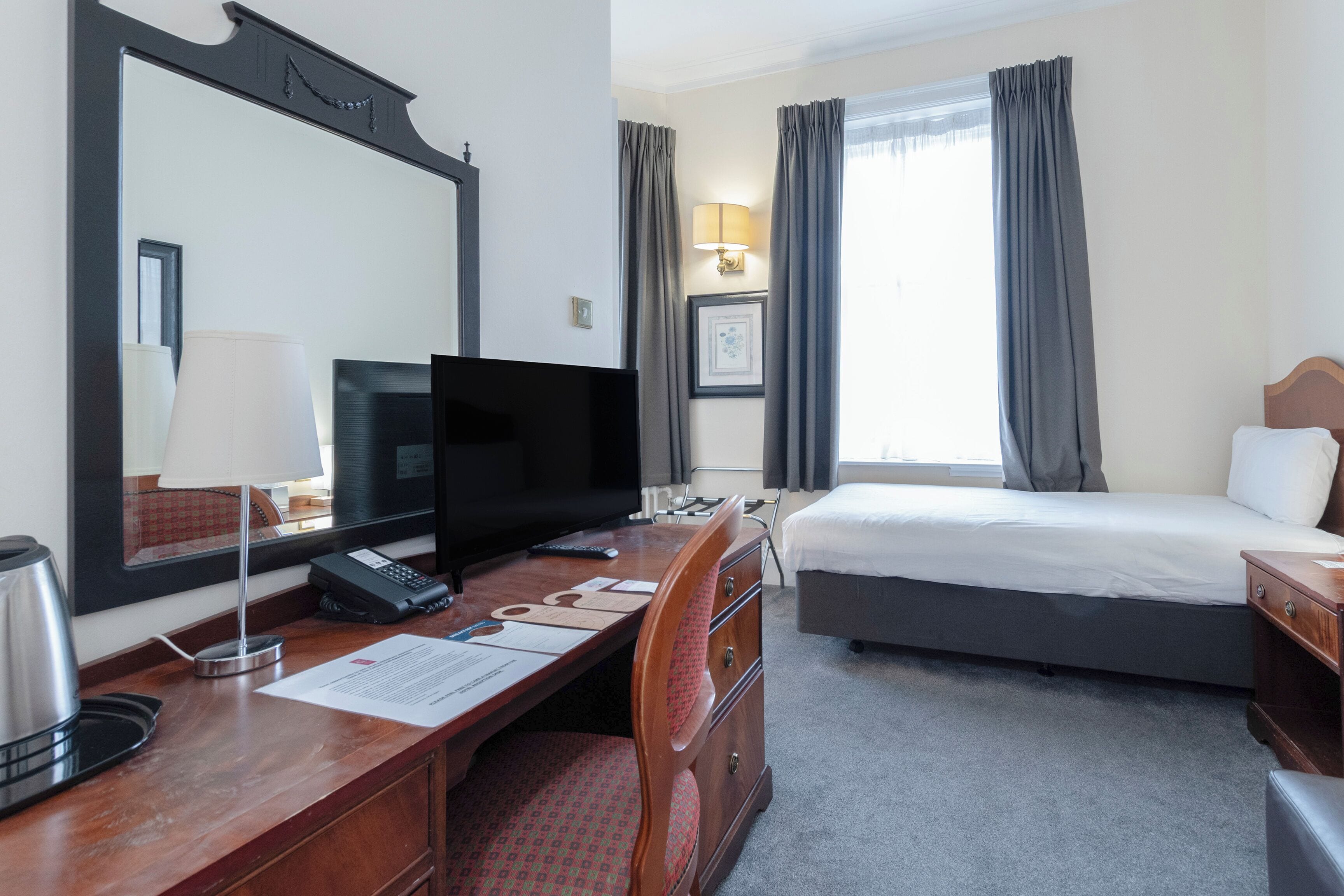 Quarto de hotel com cama, escrivaninha com televisão, cadeira, luminária e janela com cortinas.