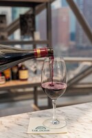Uma garrafa de vinho a servir vinho tinto num copo sobre uma mesa de mármore, com vista para a paisagem urbana ao fundo.