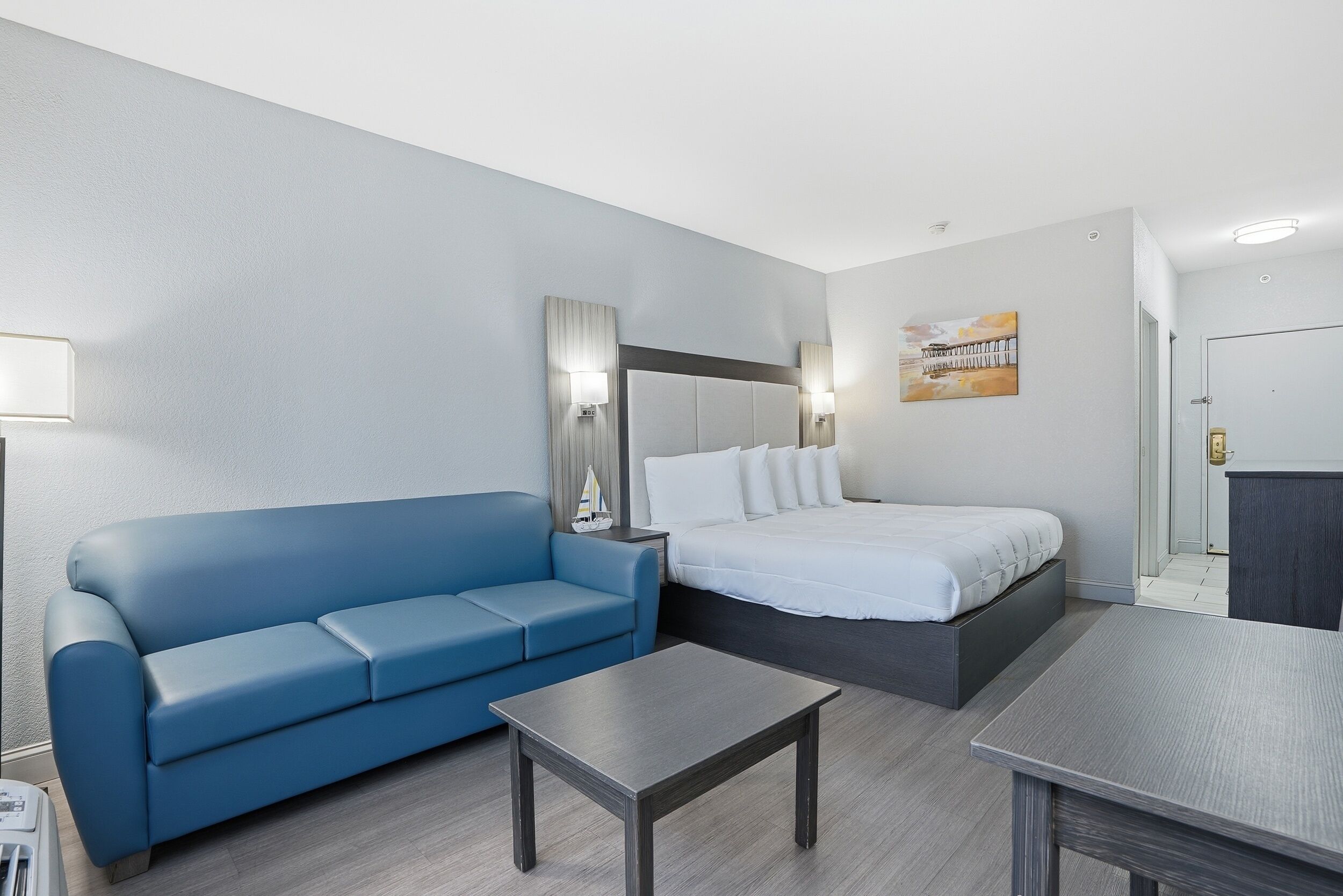 Quarto de hotel com sofá azul, cama com roupa de cama branca, uma mesinha e um quadro na parede.