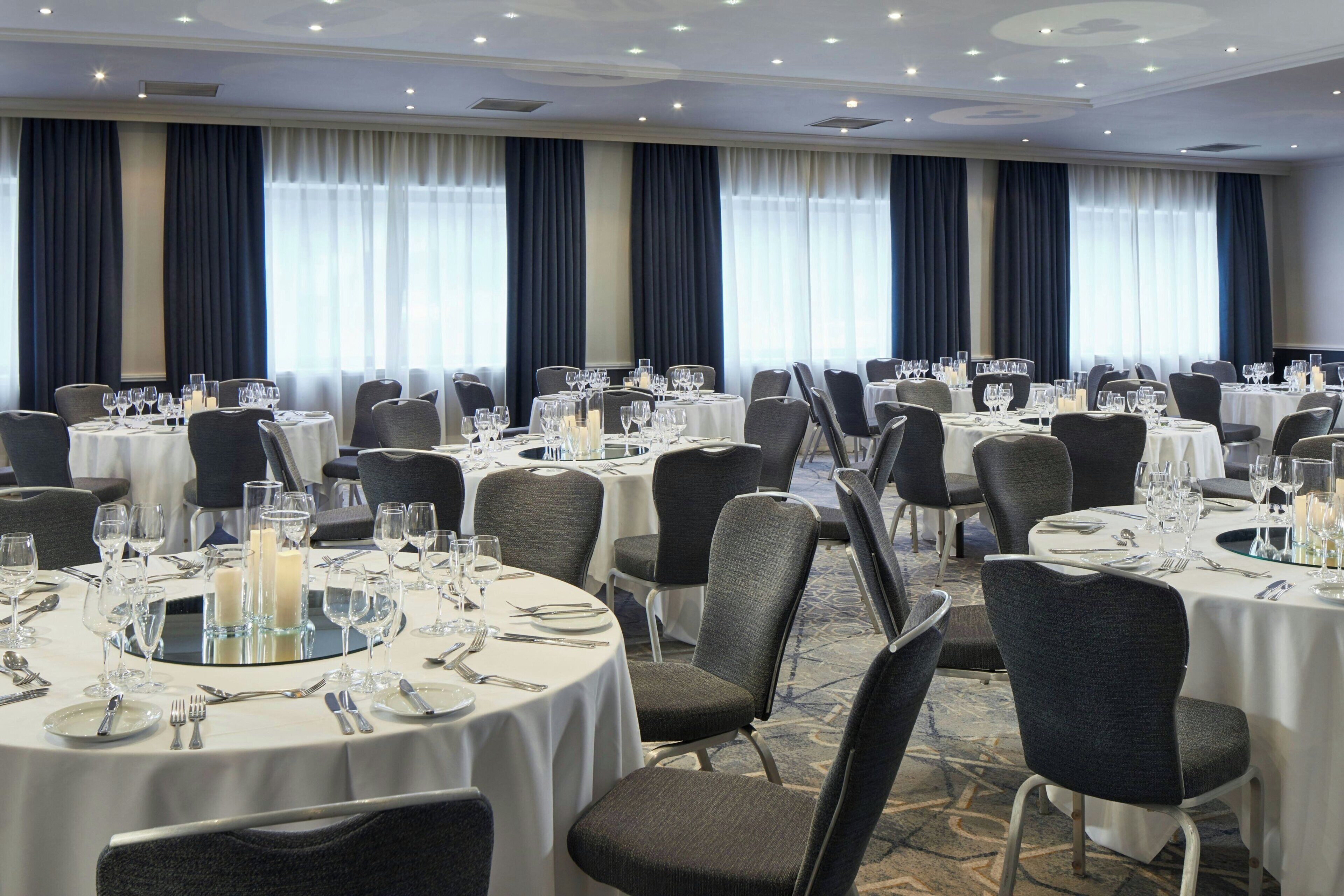Salão de banquetes com mesas redondas preparadas para refeições, com toalhas brancas, cadeiras e copos.