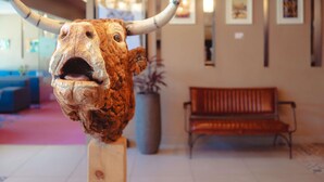 Escultura de cabeça de touro sobre um pedestal de madeira em um saguão com um banco e quadros nas paredes.
