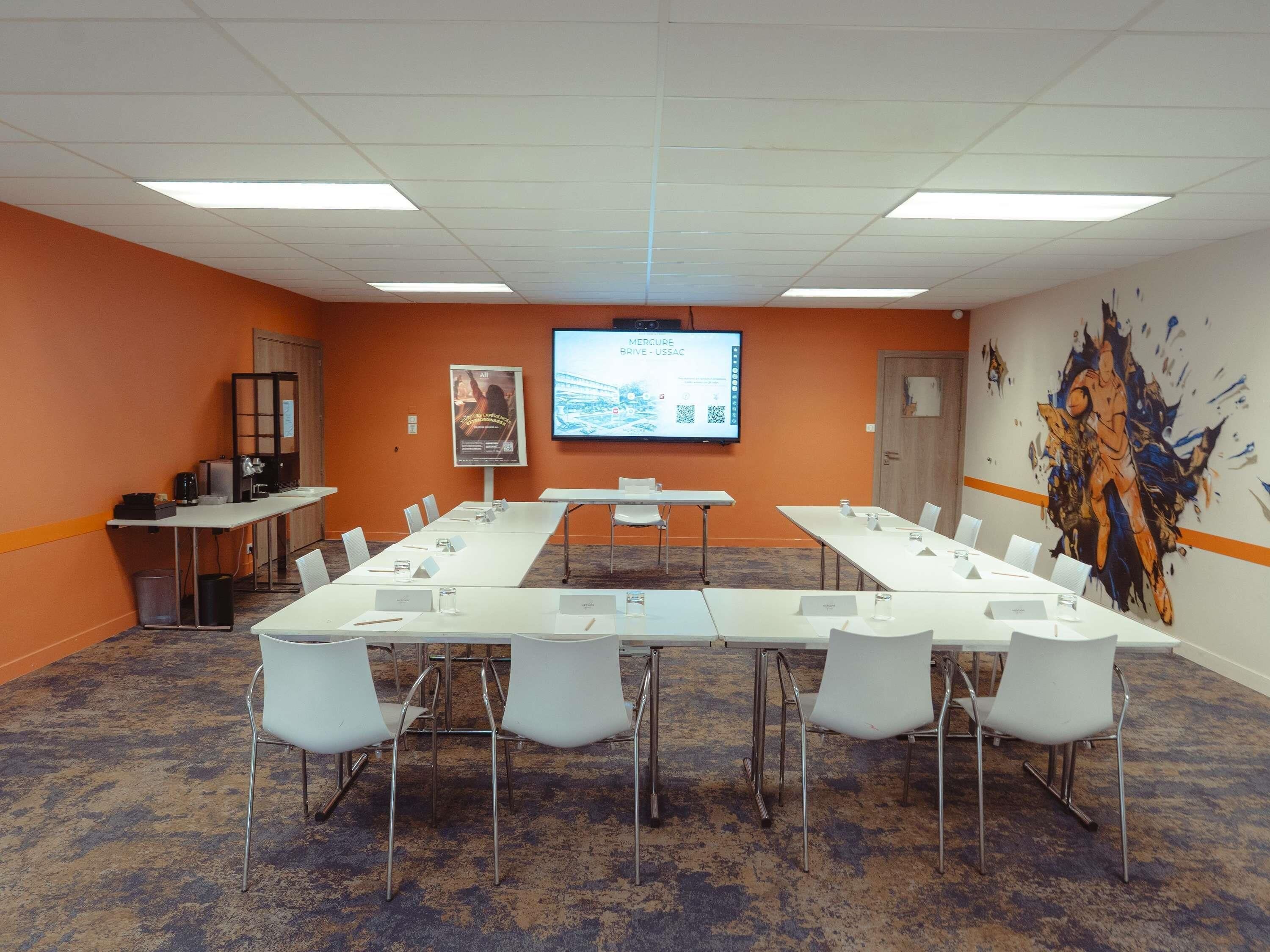 Uma sala de conferências com uma mesa comprida, cadeiras brancas, uma tela de projetor e um mural na parede.