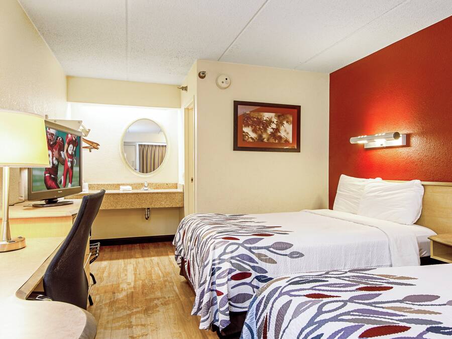 Quarto de hotel com cama, escrivaninha com computador, cadeira, espelho e luminária na parede.