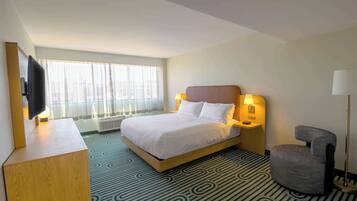 Quarto de hotel com cama, escrivaninha, cadeira, televisão e janela com cortinas.