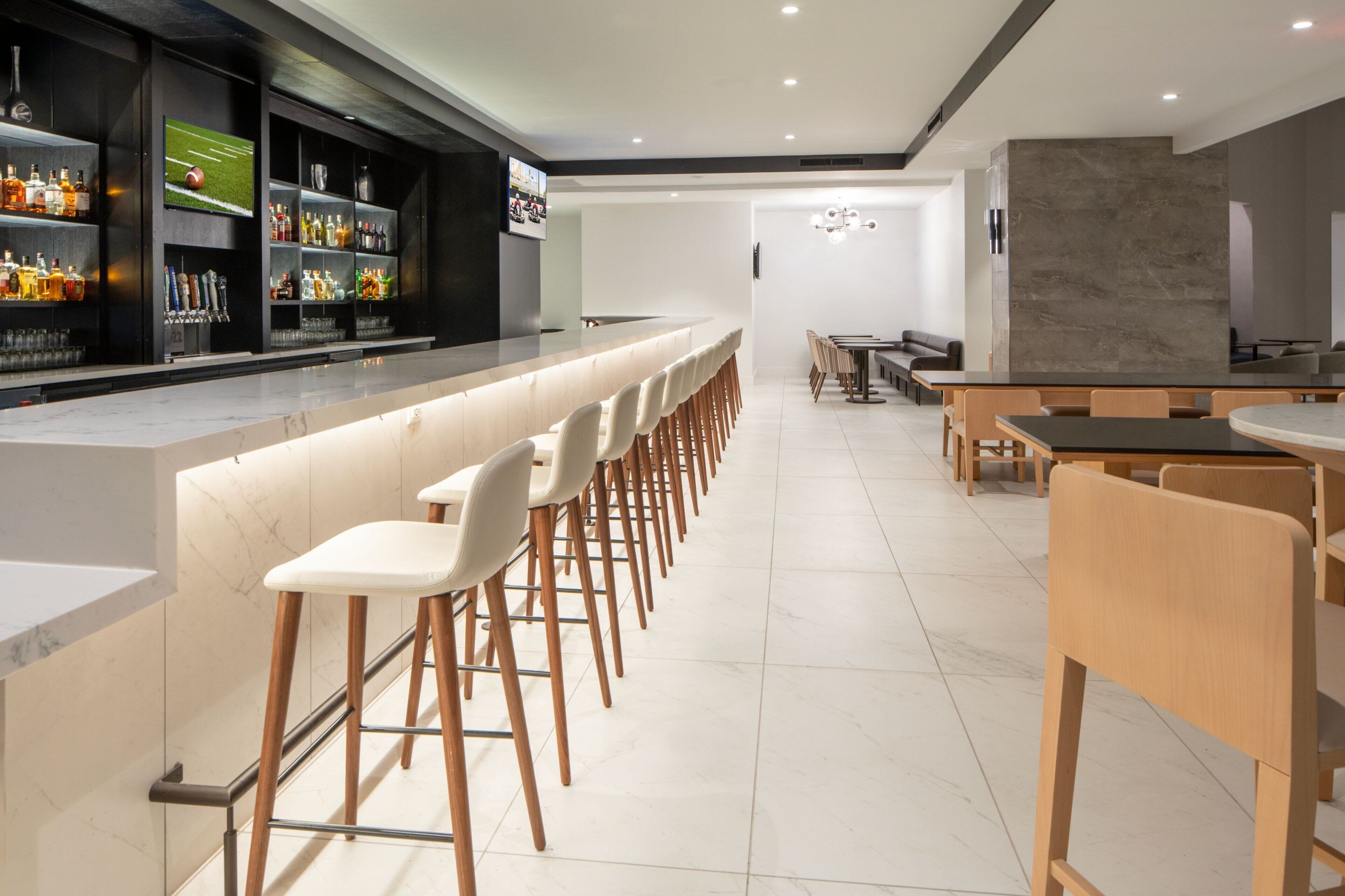Uma área de bar moderna com um balcão comprido e vários banquinhos. Há mesas e cadeiras ao fundo, indicando uma sala de jantar.