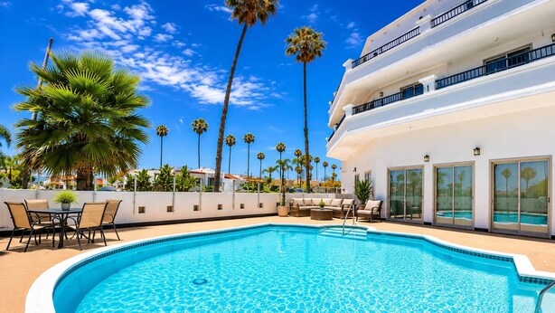 Piscina com espreguiçadeiras e palmeiras, rodeada por um edifício branco com varandas.