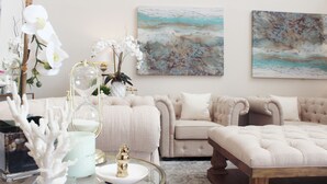 Uma sala de estar com um sofá bege, uma mesa de centro de vidro e objetos decorativos, incluindo orquídeas e um vaso de vidro.