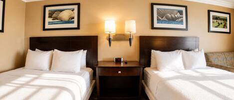 Quarto de hotel com duas camas, uma mesa de cabeceira com relógio, luminárias fixadas na parede e quadros enquadrados.