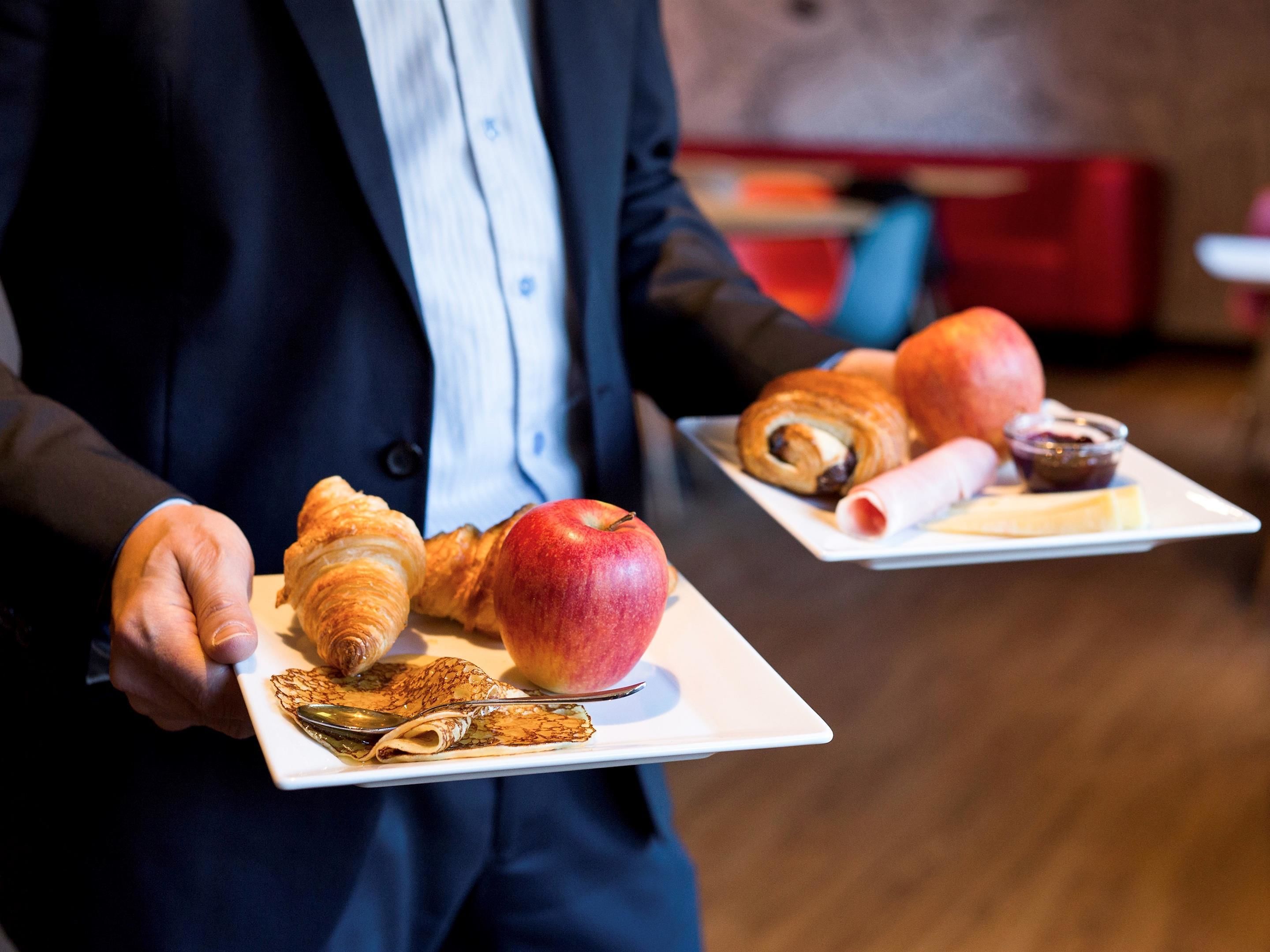 Uma pessoa segurando uma bandeja com um croissant, uma maçã e um doce com recheio de chocolate.