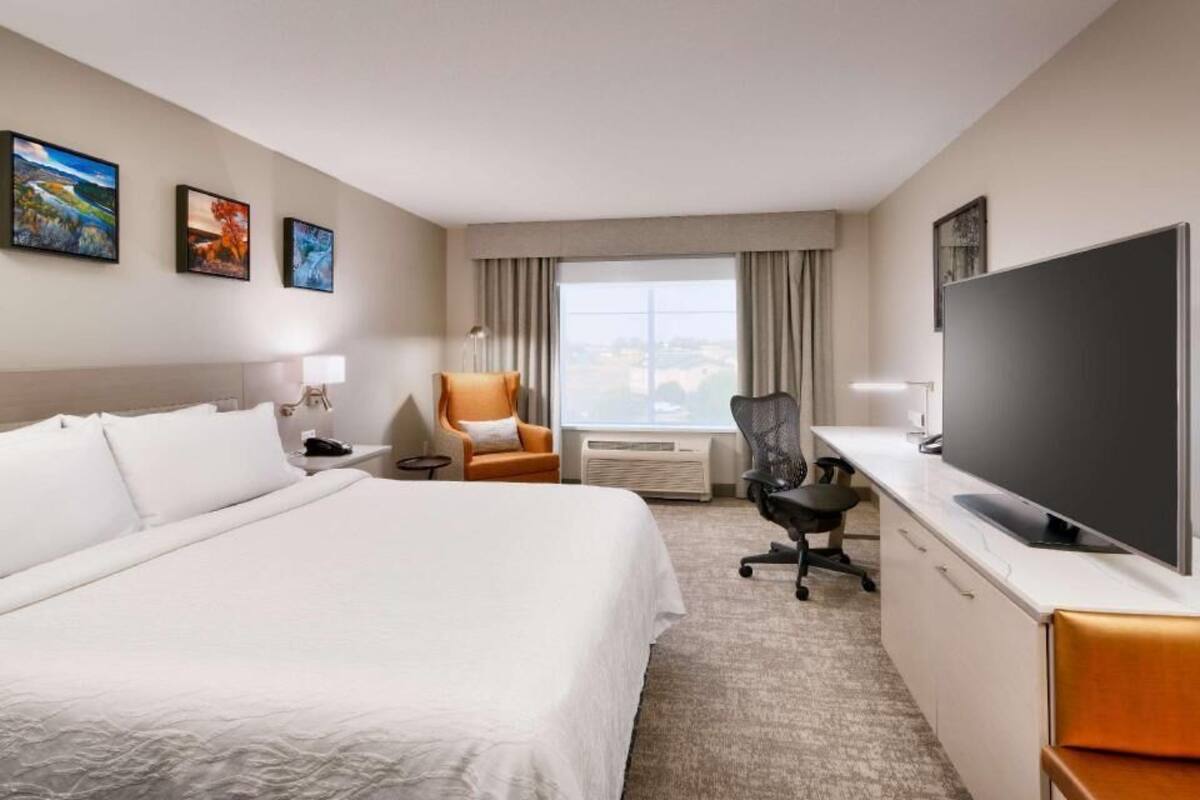 Quarto de hotel com uma cama grande, uma escrivaninha com televisão de tela plana, uma cadeira e uma janela com cortinas.