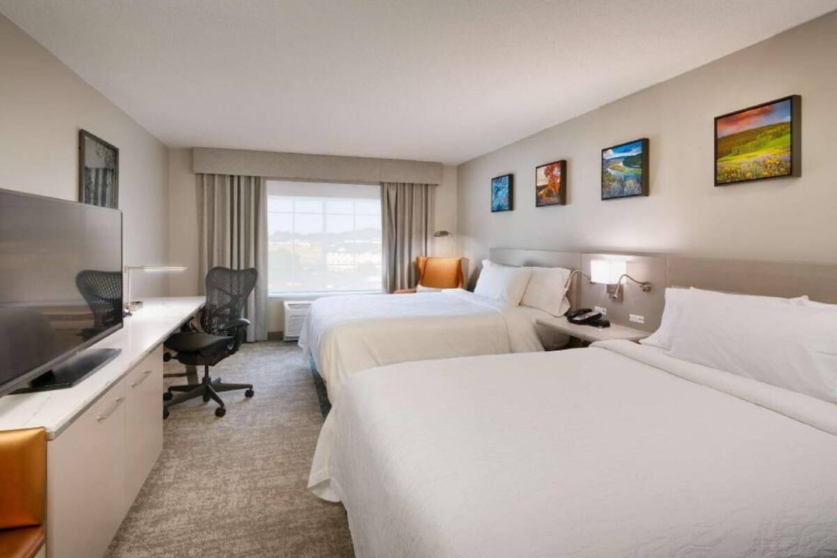 Quarto de hotel com duas camas, uma mesa de trabalho com televisão, uma cadeira e uma janela com cortinas.