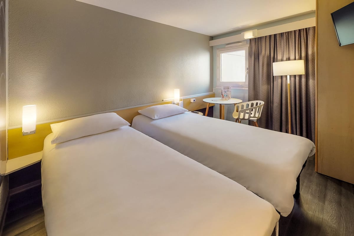 Quarto de hotel com duas camas, uma mesa pequena, uma cadeira, televisão e janela com cortinas.