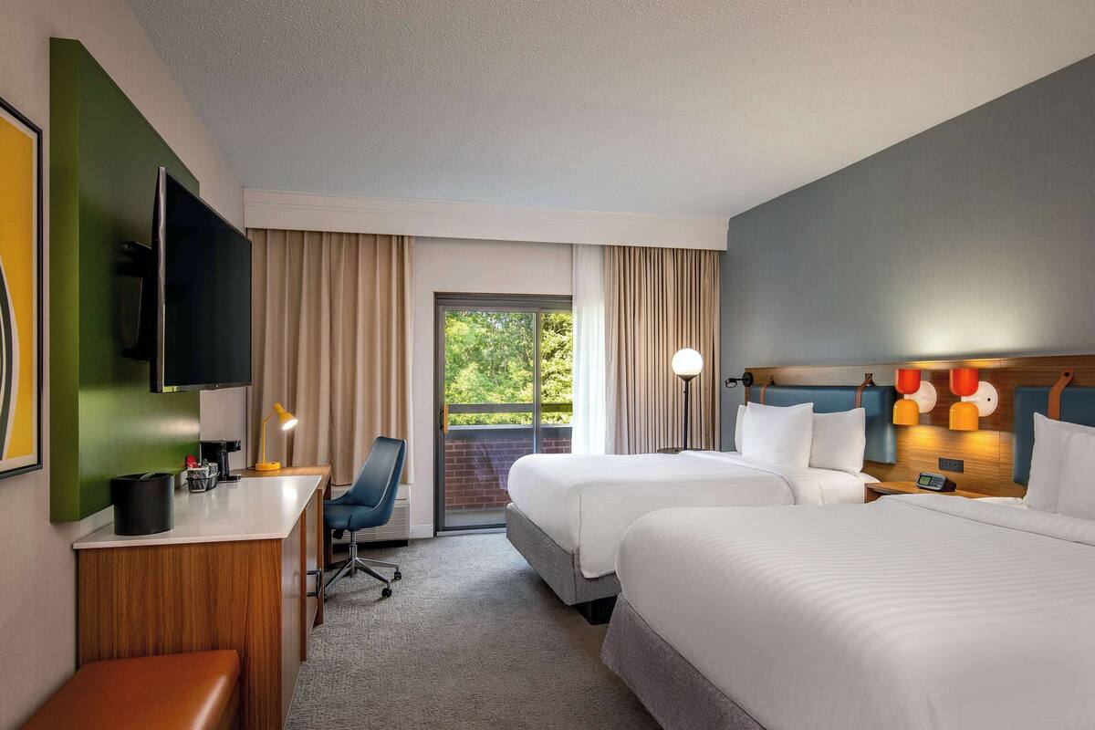 Quarto de hotel com duas camas, uma escrivaninha, uma cadeira, televisão e janela com cortinas.