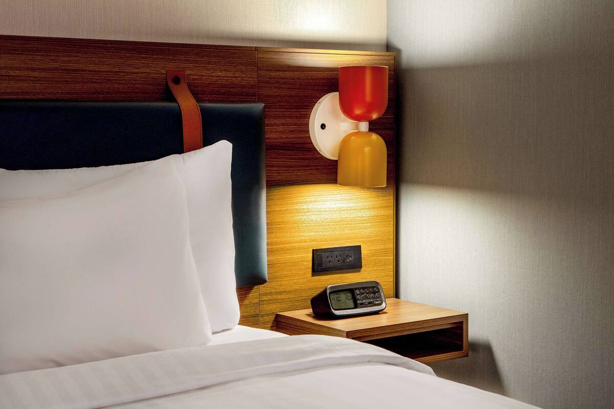 Uma cama bem arrumada com cabeceira de madeira, mesa de cabeceira com relógio e luminária na parede.