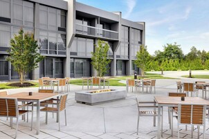 Um pátio externo com mesas e cadeiras, uma lareira e um edifício moderno com varandas e janelas amplas.