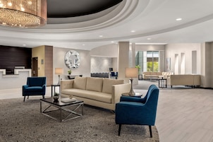 Um hall amplo com design moderno, apresentando um lustre grande, um espelho redondo e uma área de estar com um sofá bege e poltronas azuis.