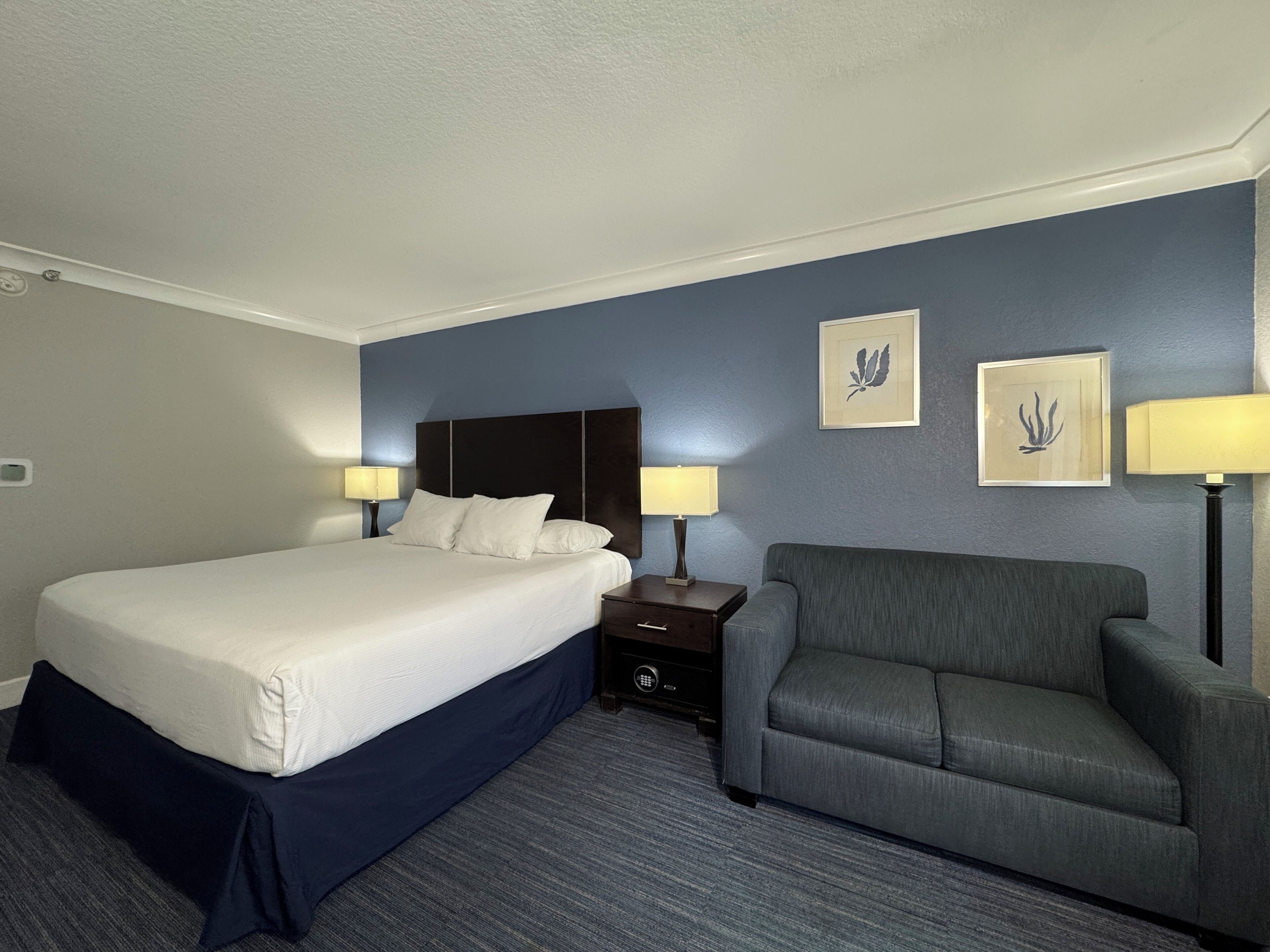 Quarto de hotel com uma cama grande, duas mesas de cabeceira com luminárias, uma poltrona cinza e duas obras de arte na parede.