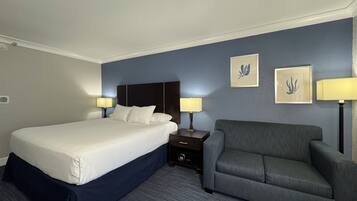 Quarto de hotel com uma cama grande, duas mesas de cabeceira com luminárias, uma poltrona cinza e duas obras de arte na parede.