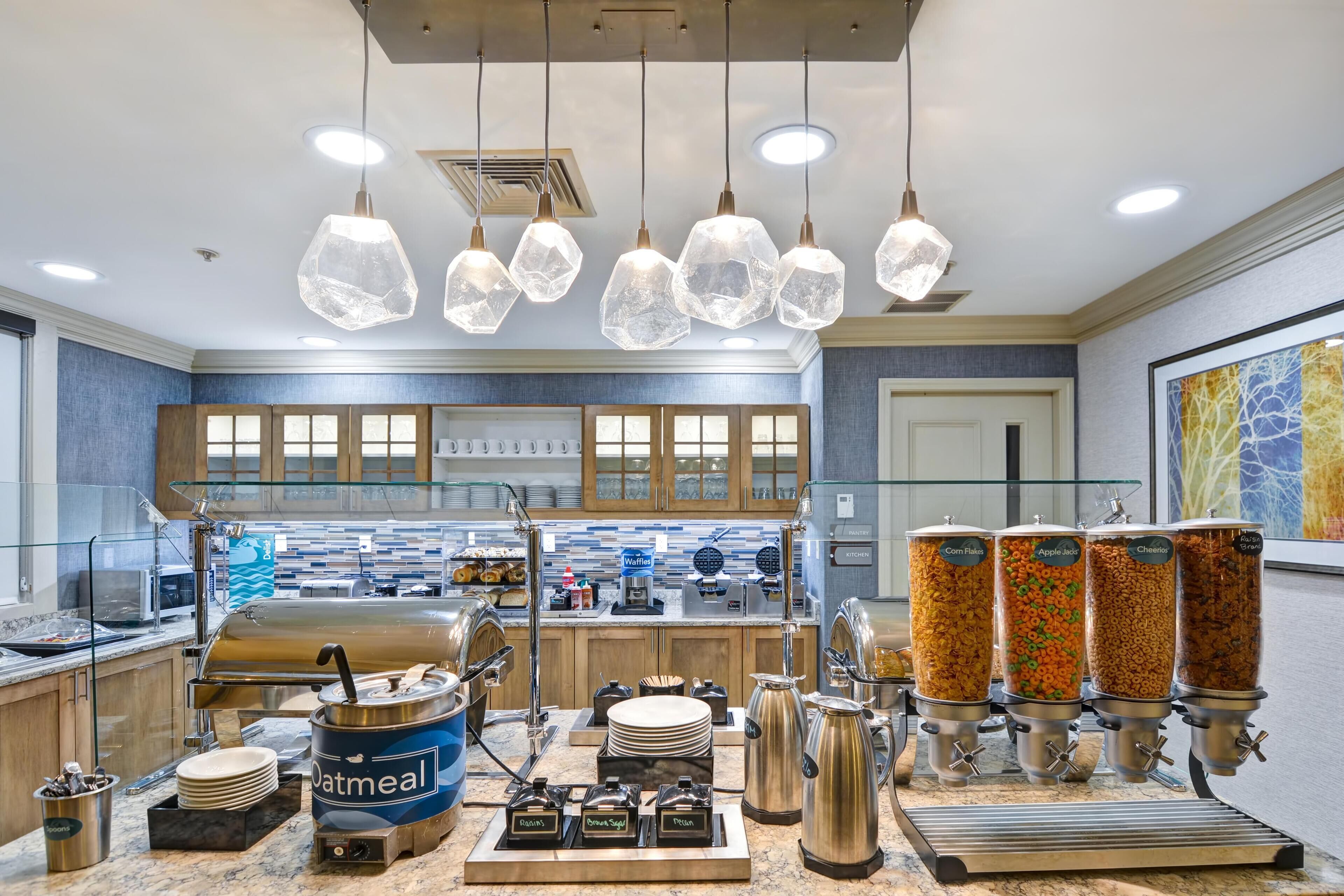 Disposição de buffet com diversos distribuidores de alimentos, um espaço para café e um balcão com pratos e talheres.