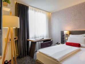 Quarto de hotel com cama, mesa de cabeceira, luminária, cadeira e escrivaninha com cadeira.