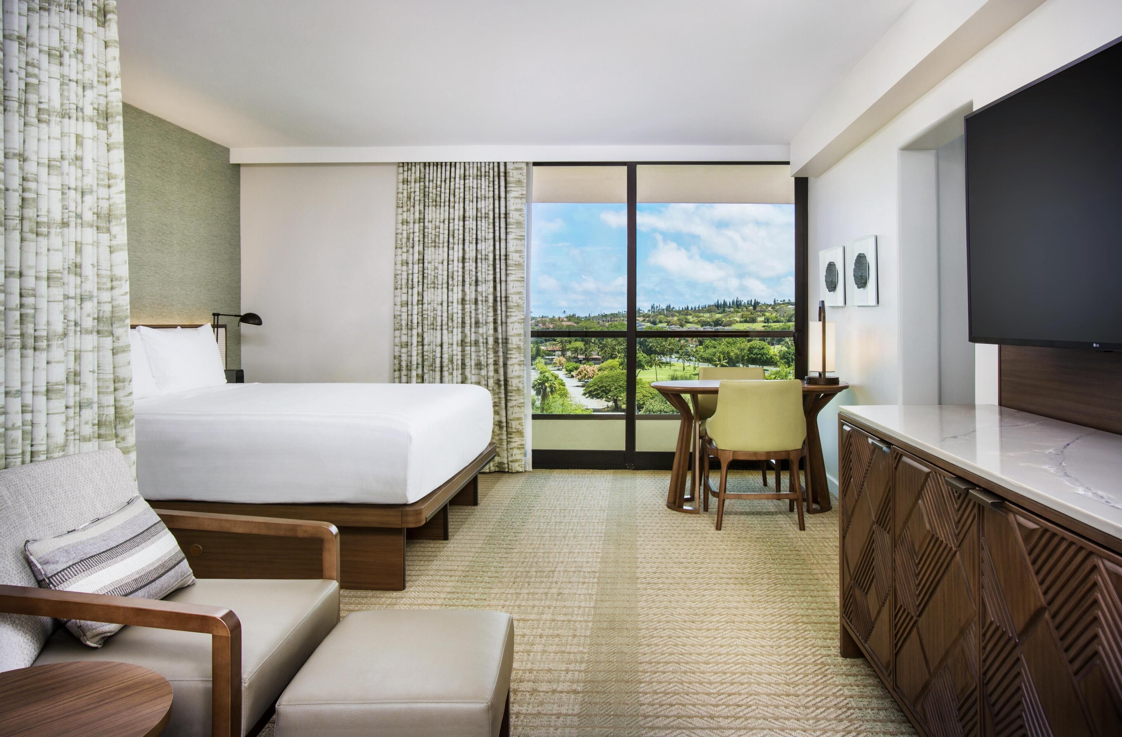 Quarto de hotel com cama, escrivaninha, cadeira, televisão e vista para uma paisagem verde.