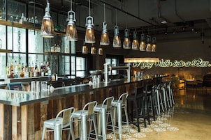 Uma área de bar com um balcão comprido, banquetas de metal, luminárias pendentes e diversas garrafas em prateleiras.