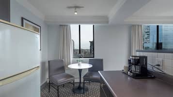 Quarto de hotel moderno com cozinha compacta, área de refeições com duas cadeiras e uma mesa pequena, e uma janela grande com vista para a cidade.