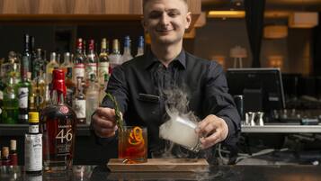 Um bar com um bartender preparando uma bebida, uma garrafa de uísque Maker's 46, e diversas outras garrafas ao fundo.