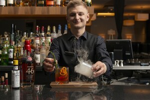 Um bar com um bartender preparando uma bebida, uma garrafa de uísque Maker's 46, e diversas outras garrafas ao fundo.