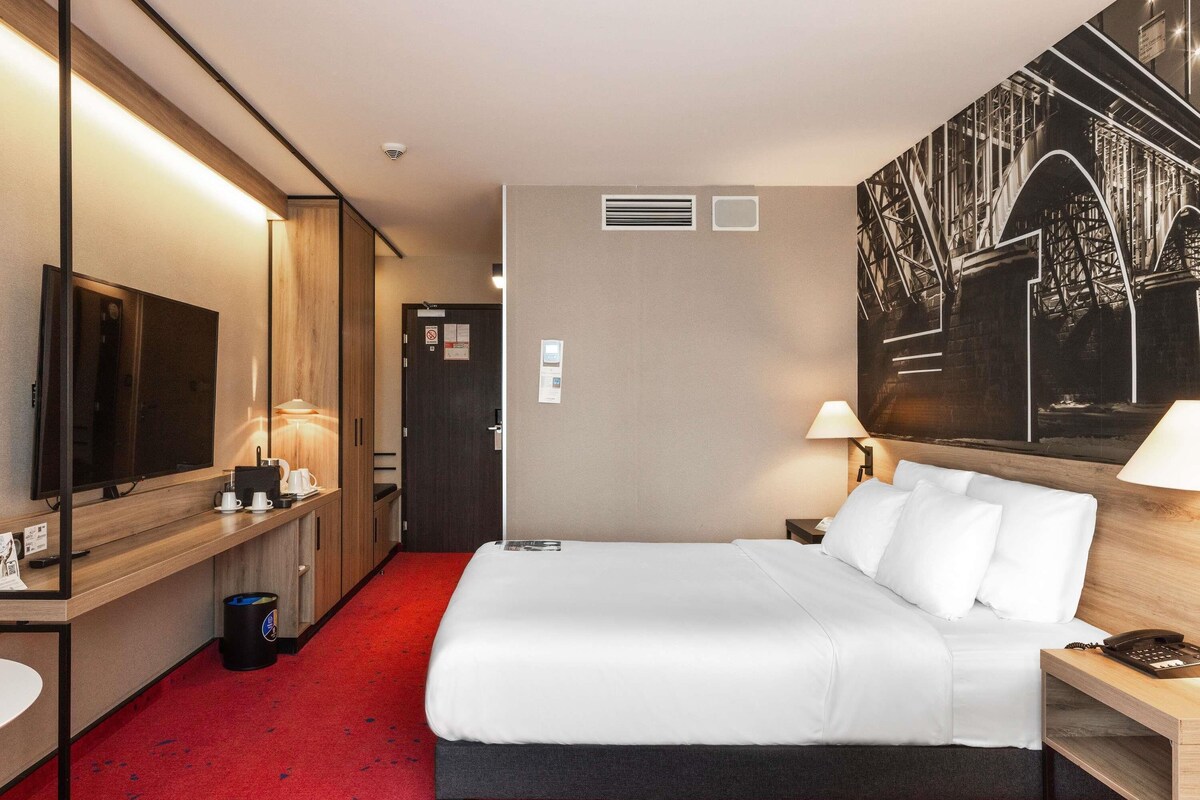 Quarto de hotel com cama, televisão, escrivaninha com computador, espelho e um quadro na parede.