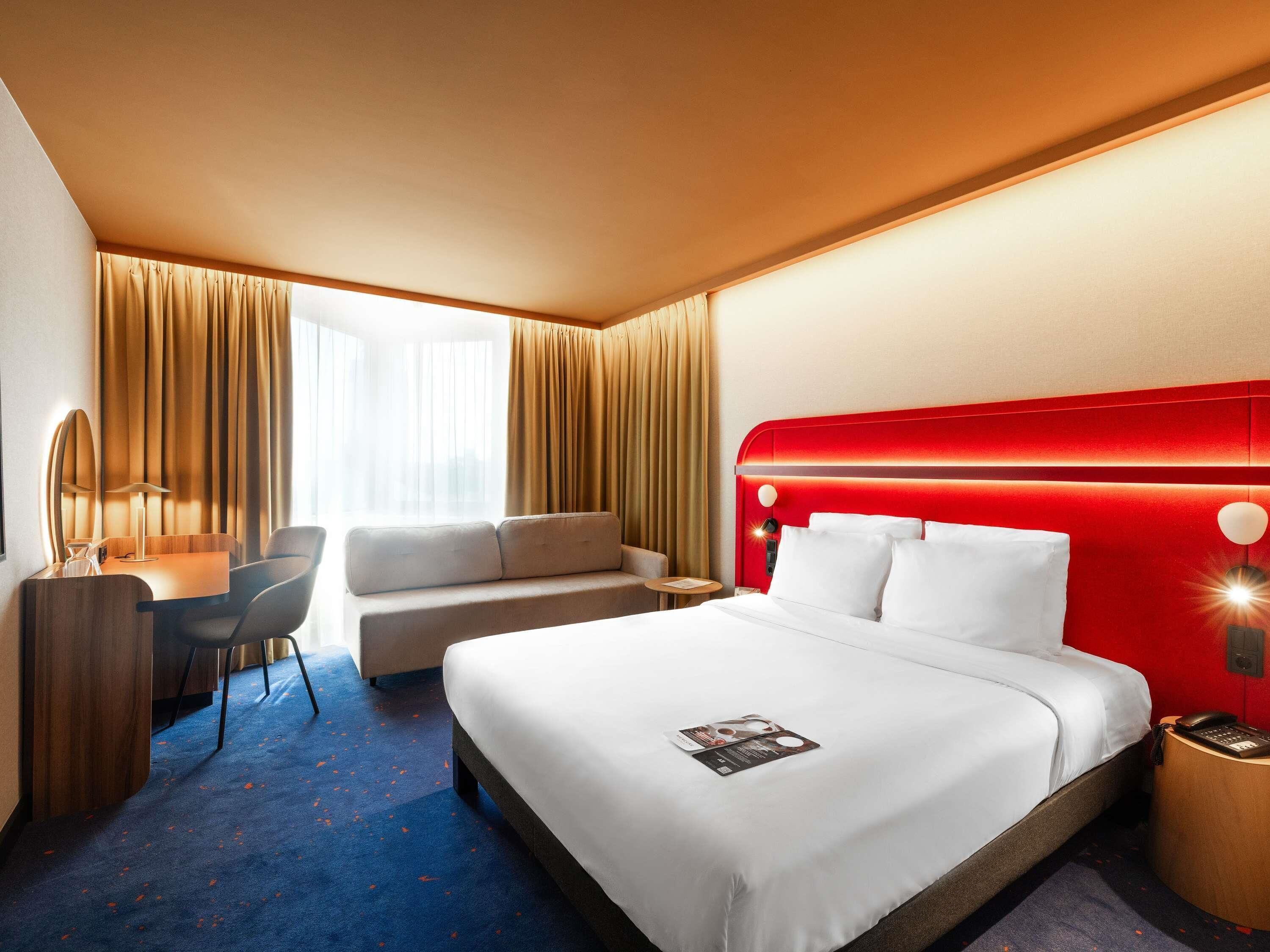 Quarto de hotel com cama, escrivaninha, cadeira, sofá e janela com cortinas.