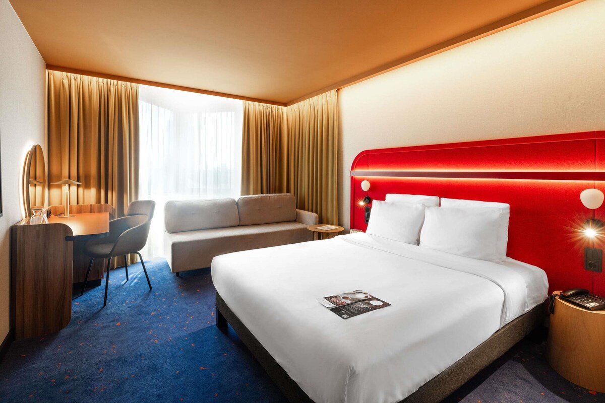 Quarto de hotel com cama, escrivaninha, cadeira, sofá e janela com cortinas.