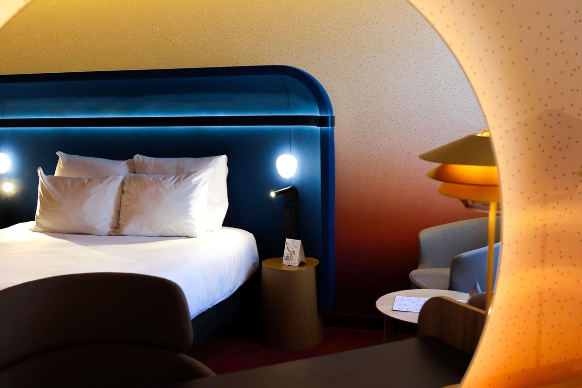 Quarto de hotel com cama, mesa de cabeceira, abajur e uma pequena mesa com uma cadeira.