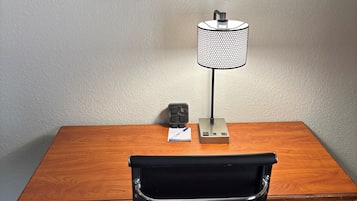 Uma escrivaninha de madeira com uma cadeira preta, uma luminária, um filtro de linha e um quadro na parede.