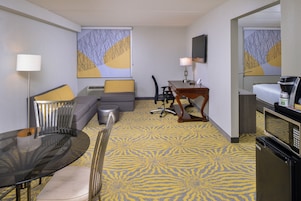 Quarto de hotel com mesa de vidro para refeições, sofá com almofadas, escrivaninha com abajur, micro-ondas e cafeteira.