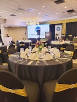 Sala de conferências com mesas redondas preparadas para um evento formal, com cartões de nome, copos de água e arranjos florais.